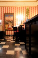 Um hall de hotel com piso xadrez, papel de parede listrado, um pôster emoldurado de um horizonte urbano, um vaso com flores e um abajur.