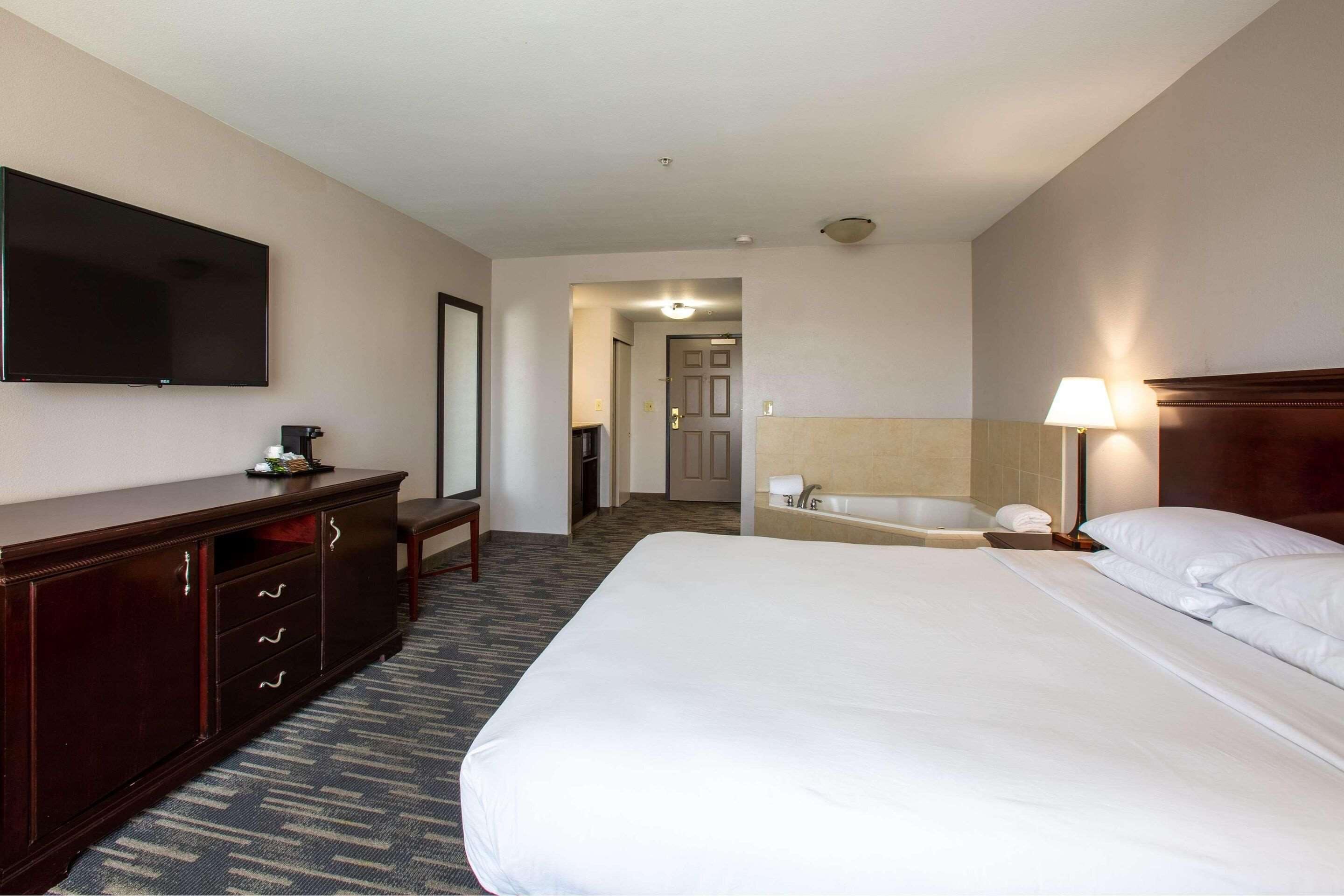 Quarto de hotel com uma cama grande, televisão de tela plana na parede, cómoda de madeira, banheiro com banheira e uma luminária na mesa de cabeceira.