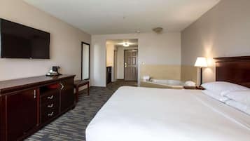 Quarto de hotel com uma cama grande, televisão de tela plana na parede, cómoda de madeira, banheiro com banheira e uma luminária na mesa de cabeceira.