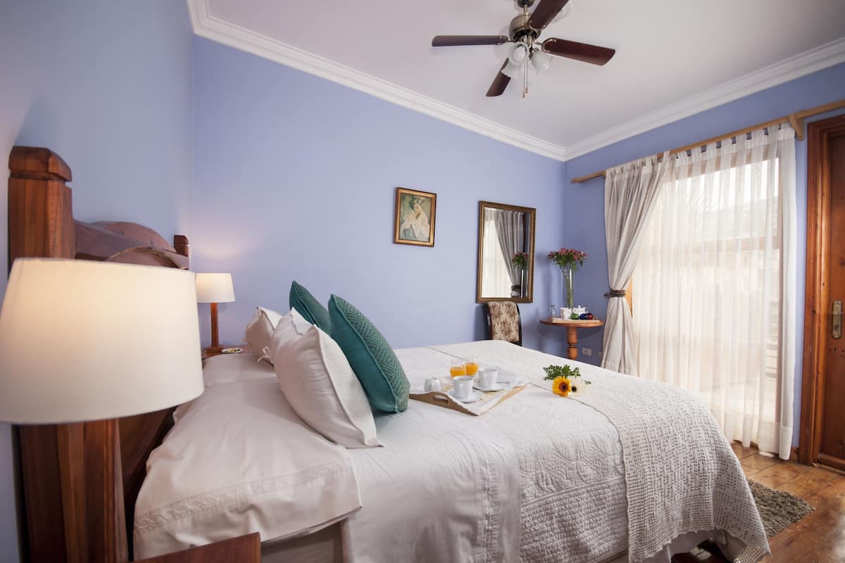 Um quarto com cama, mesa de cabeceira, abajur, ventilador de teto e janela com cortinas.