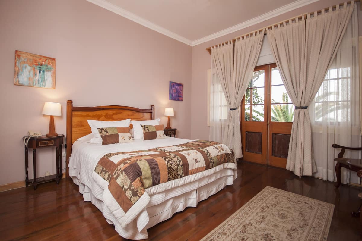 Uma cama bem arrumada com cabeceira de madeira, duas mesas de cabeceira com abajures e uma janela grande com cortinas.