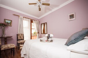 Quarto com cama, cadeira, mesa de cabeceira com um vaso, cortinas, ventilador de teto e um quadro na parede.