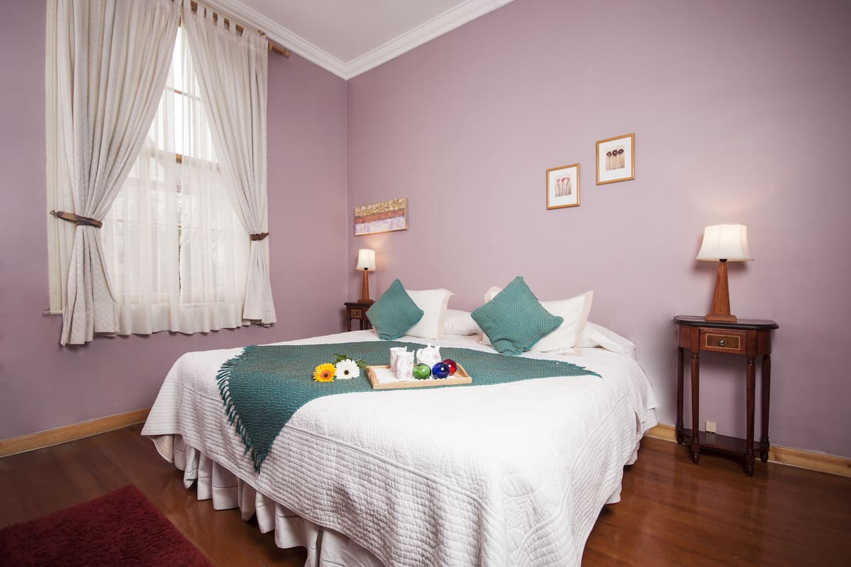 Cama bem arrumada com edredom branco e almofadas verdes, mesa de cabeceira com abajur e janela com cortinas leves.