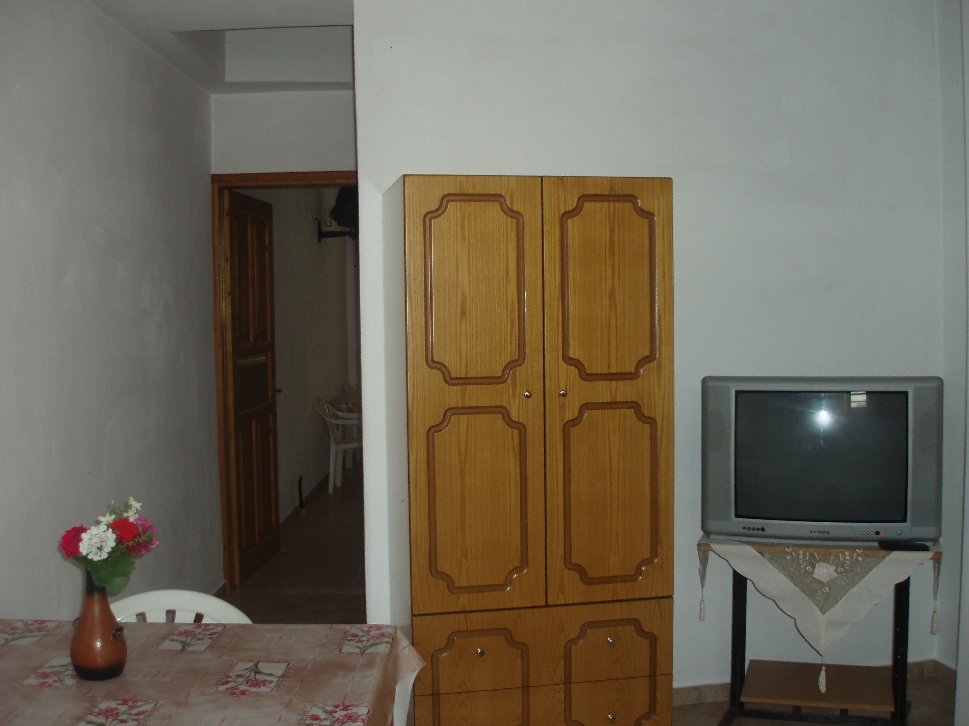 Um quarto com um guarda-roupa de madeira, uma televisão em um móvel e uma mesa com um vaso de flores.