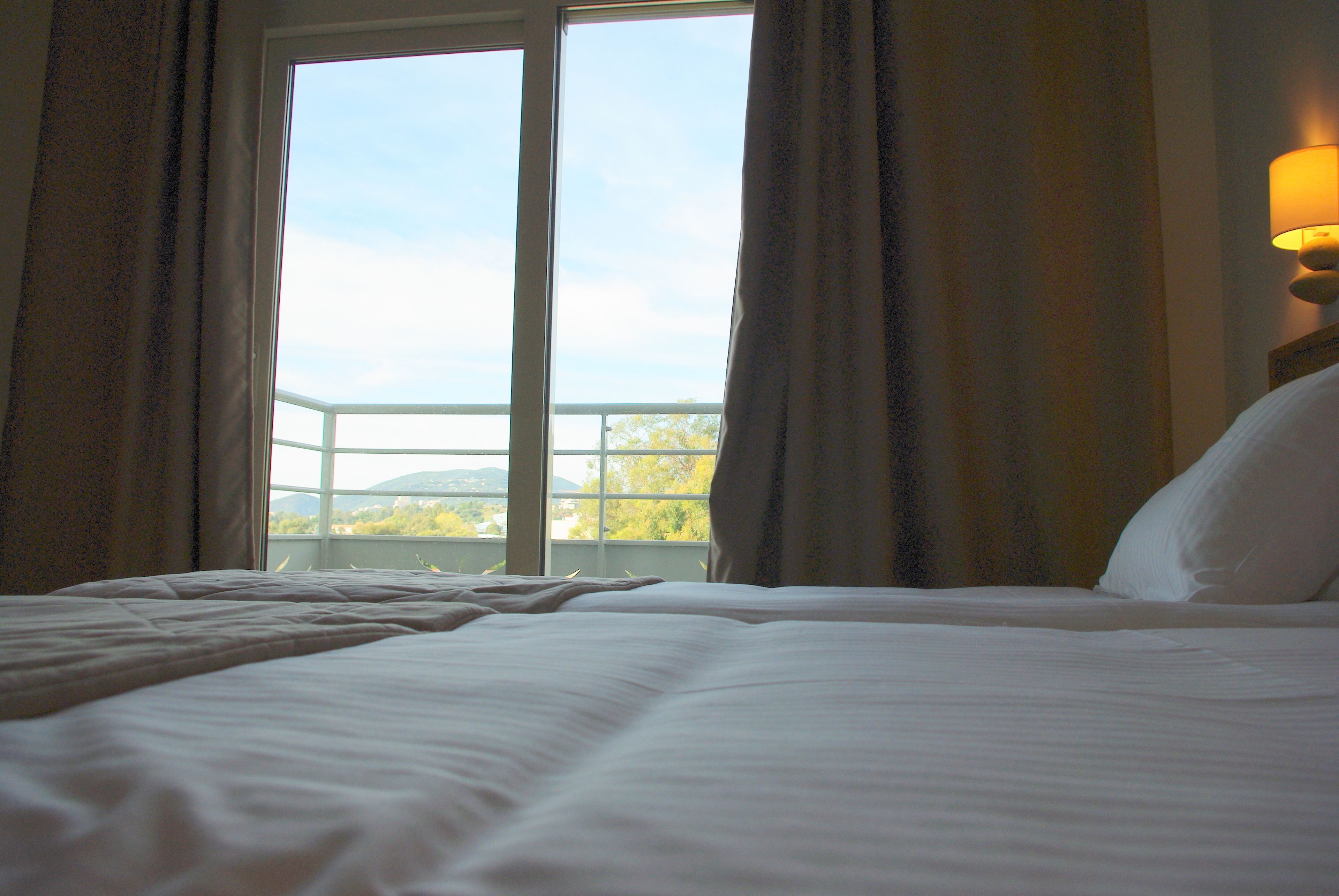 Quarto de hotel com cama, janela com cortinas e uma luminária.