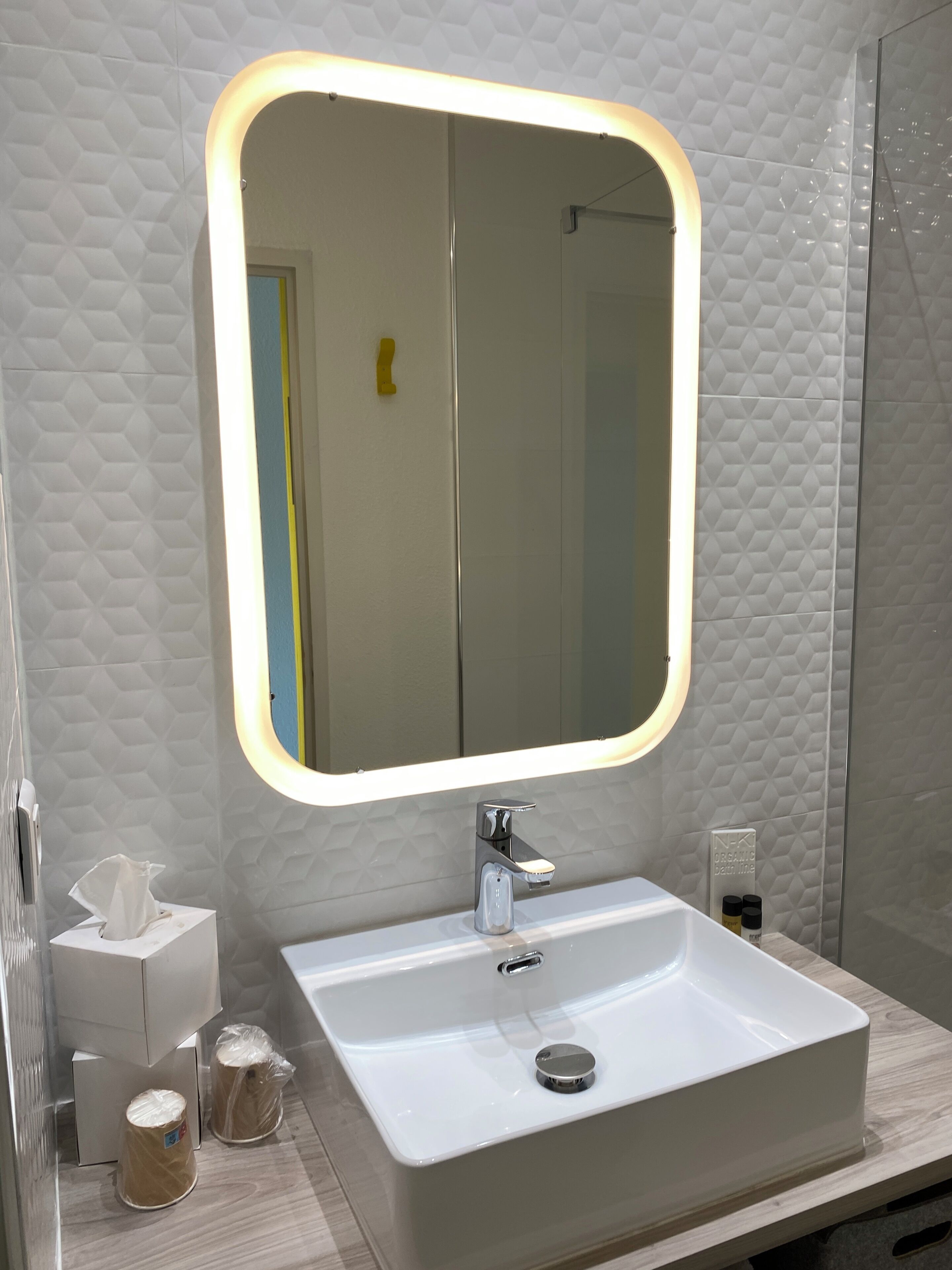 Casa de banho com lavatório branco, espelho iluminado e dispensador de toalhas.