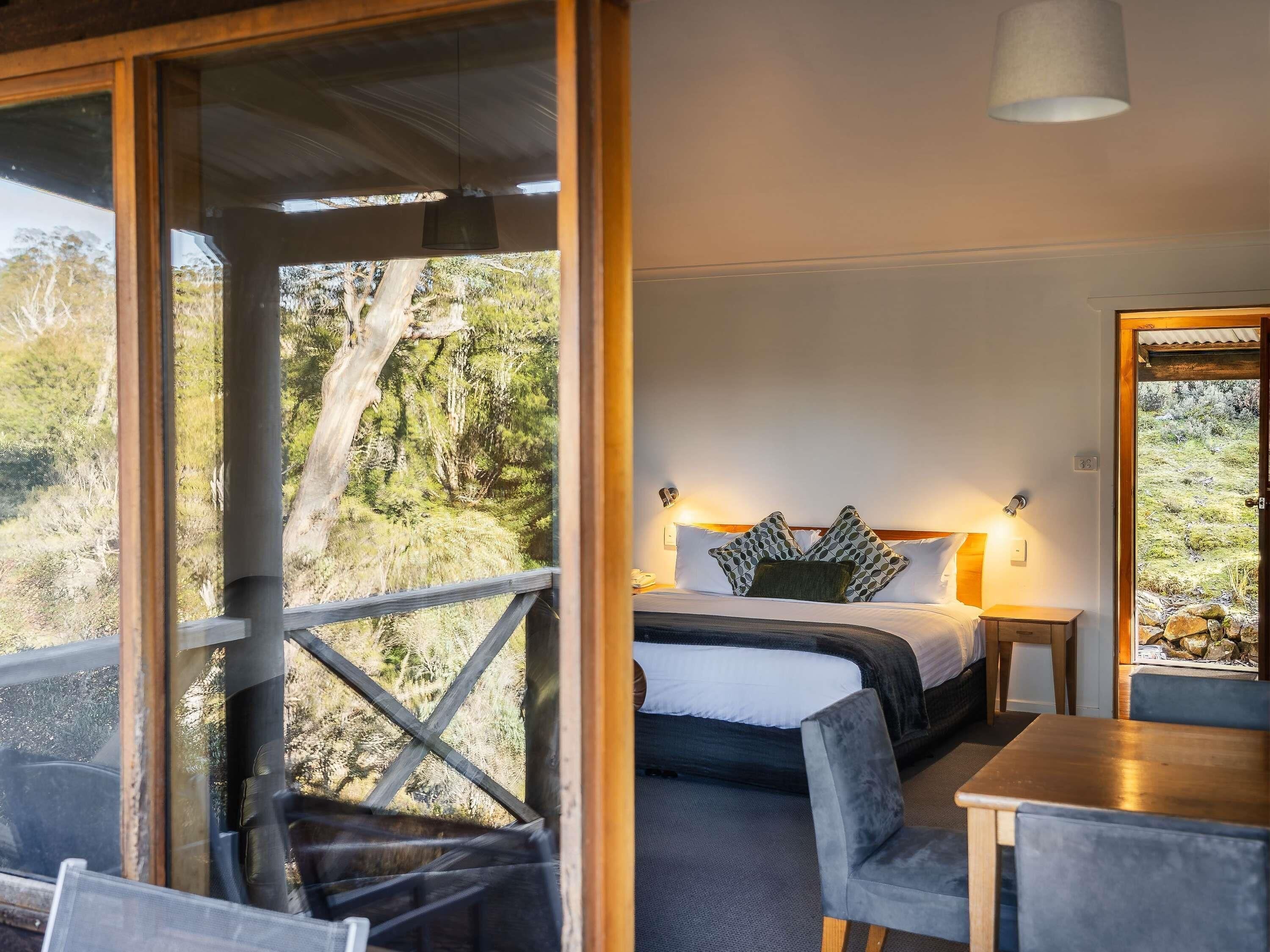 Quarto com cama, mesas de cabeceira, escrivaninha e vista para árvores pela janela.