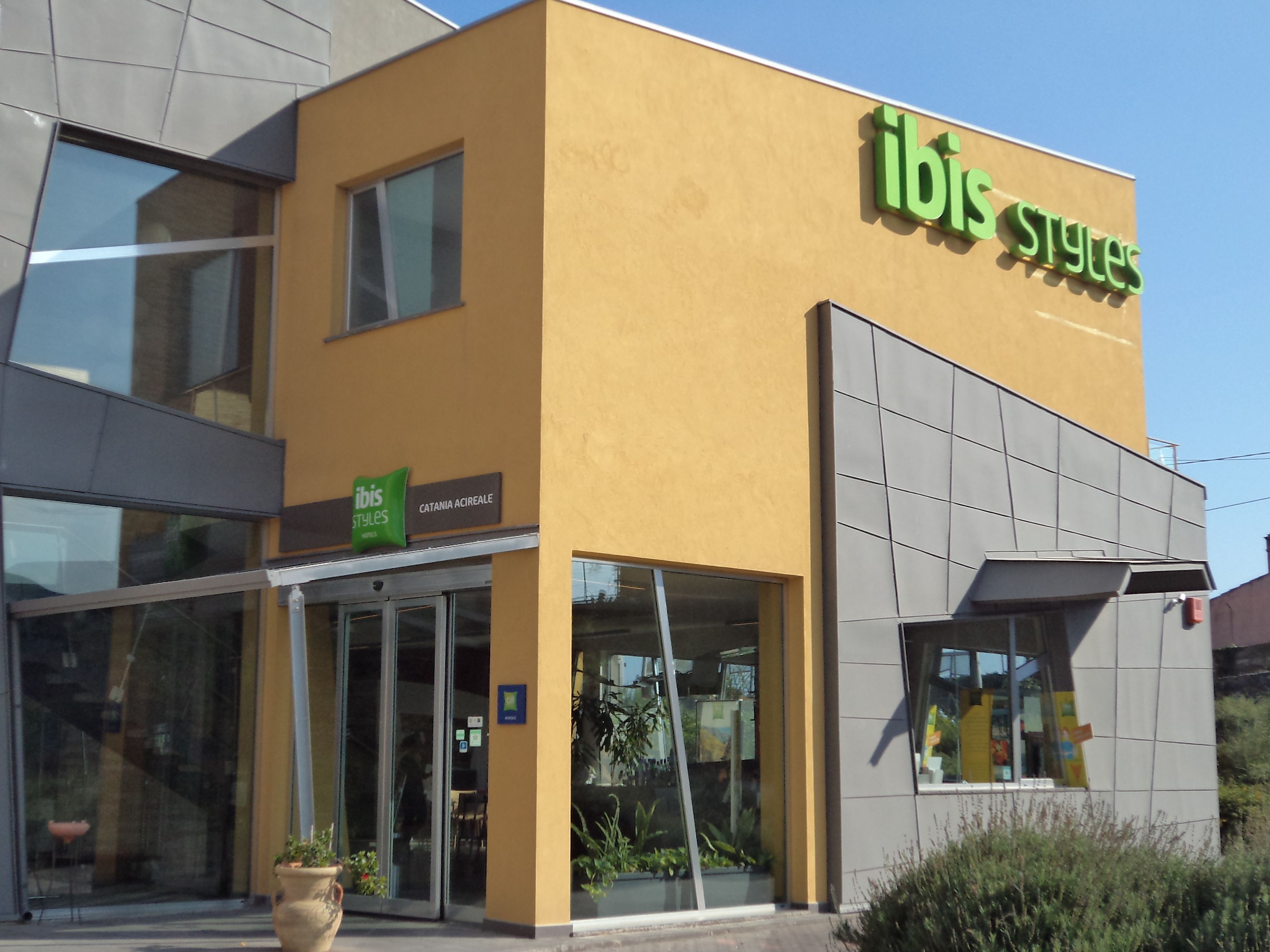 Um edifício moderno com fachada amarela e uma placa verde com a inscrição 'ibis Styles'.