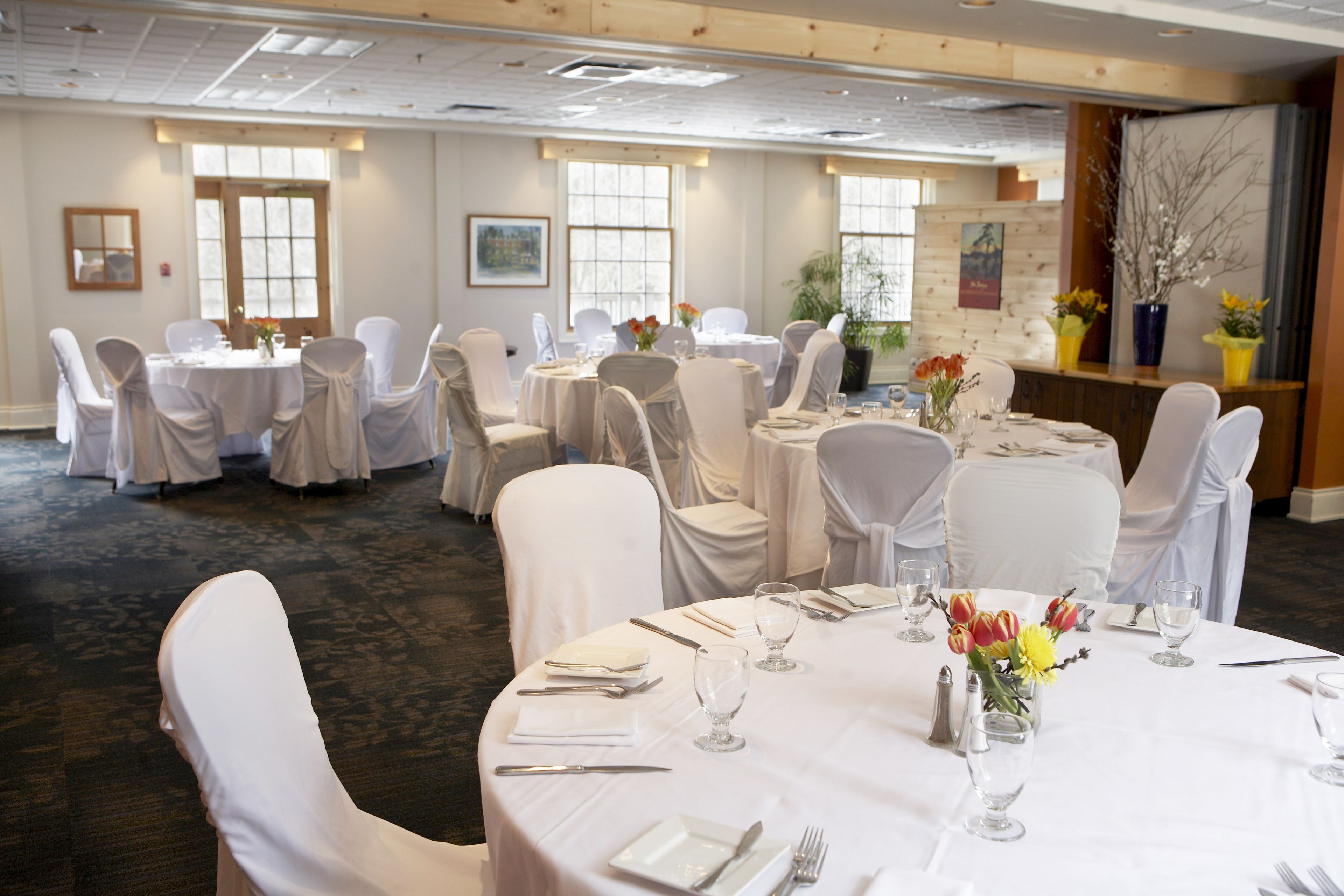 Uma sala de banquetes com mesas redondas preparadas para refeições, cadeiras brancas e centros de mesa florais.