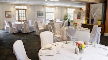Uma sala de banquetes com mesas redondas preparadas para refeições, cadeiras brancas e centros de mesa florais.