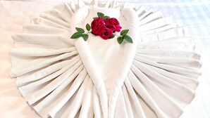 Cama bem arrumada com edredom branco e almofadas, colcha branca e arranjo decorativo de toalhas brancas com rosas vermelhas.