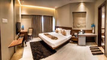 Quarto de hotel moderno com cama grande, mesa de cabeceira, escrivaninha, cadeira, pequeno espaço com cozinha e banheiro.