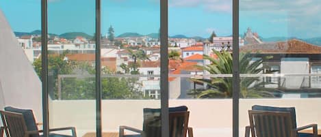 Varanda com três cadeiras de madeira e uma mesa pequena, com vista para uma paisagem urbana com edifícios de telhado vermelho e céu claro.