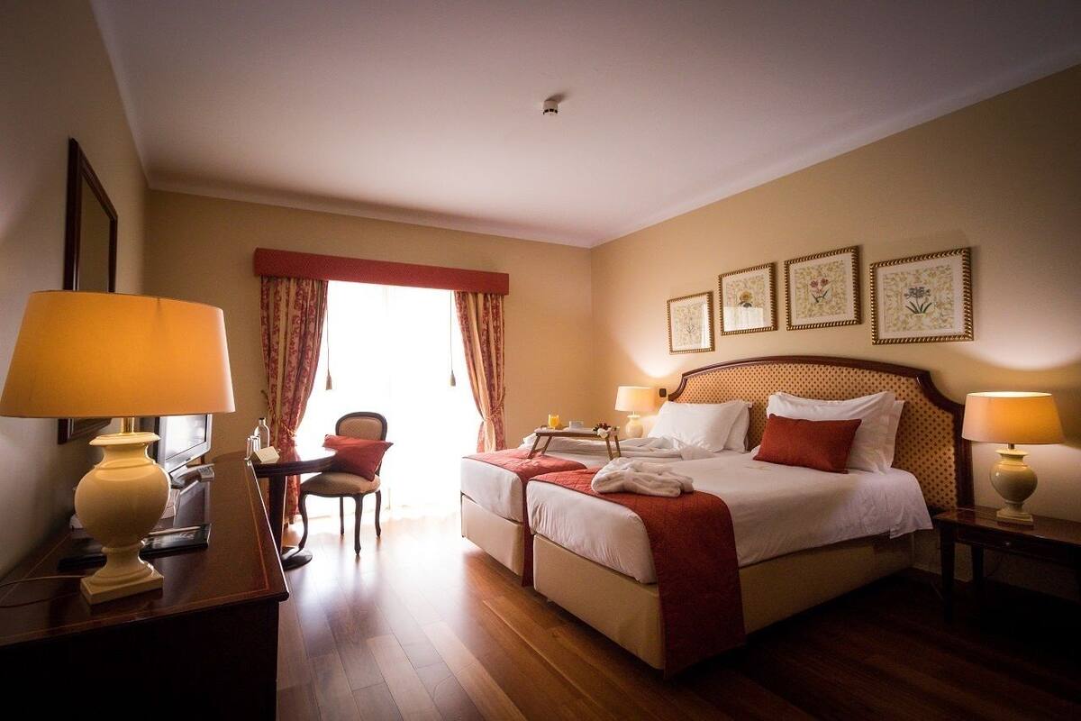 Quarto de hotel com uma cama grande, duas mesas de cabeceira com abajures, uma escrivaninha com cadeira e uma janela com cortinas.