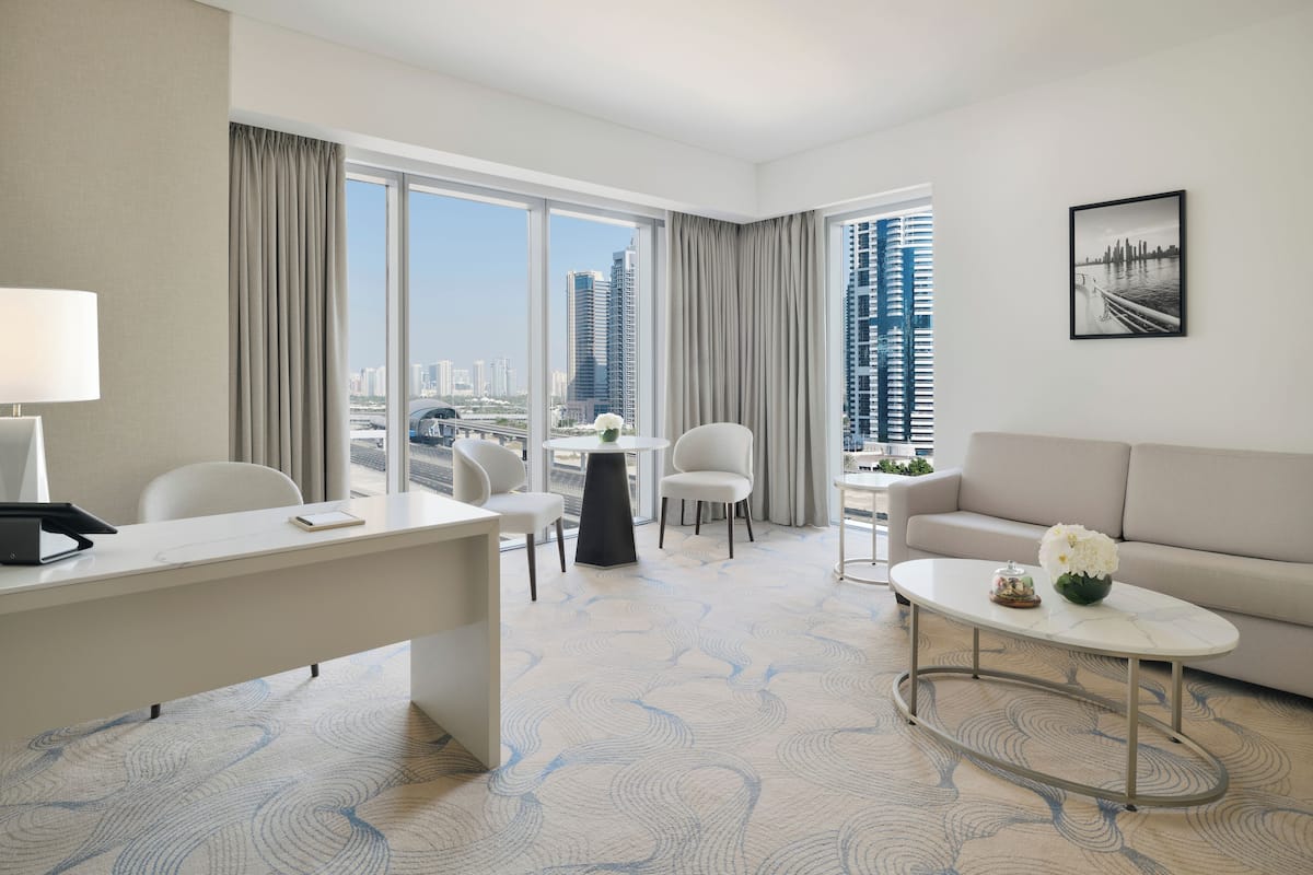 Quarto de hotel moderno com escrivaninha, duas cadeiras, um sofá e uma mesa de centro. Janelas amplas oferecem vista para a cidade.