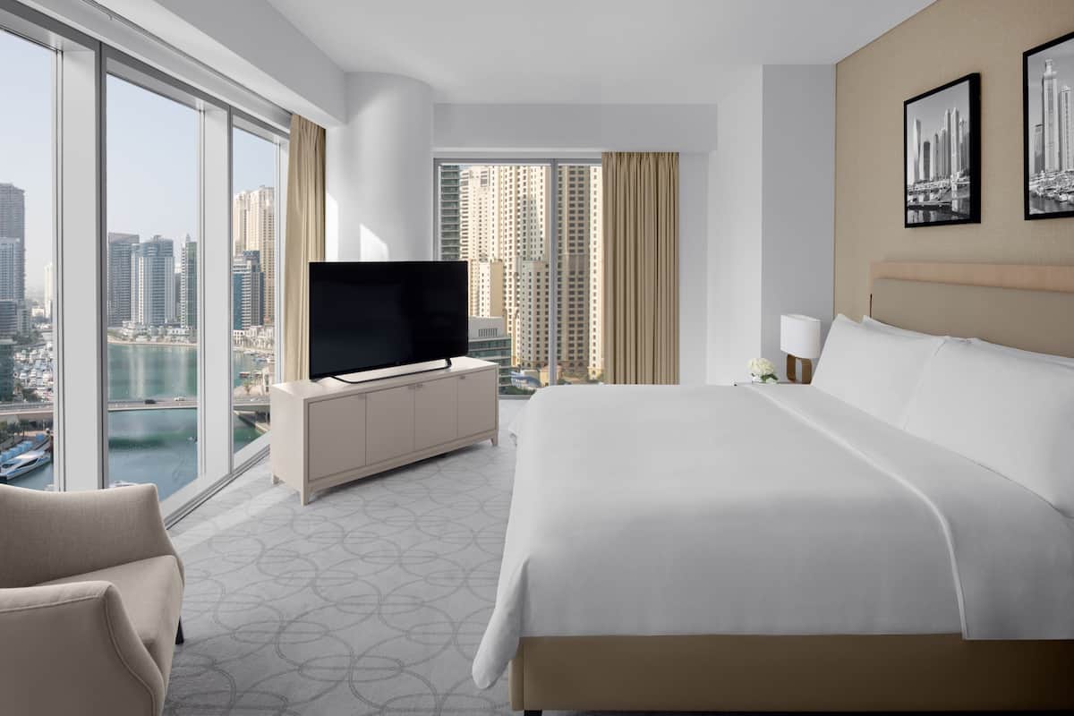 Quarto de hotel moderno com uma cama grande, televisão em um móvel, vista para a cidade e cortinas na janela.
