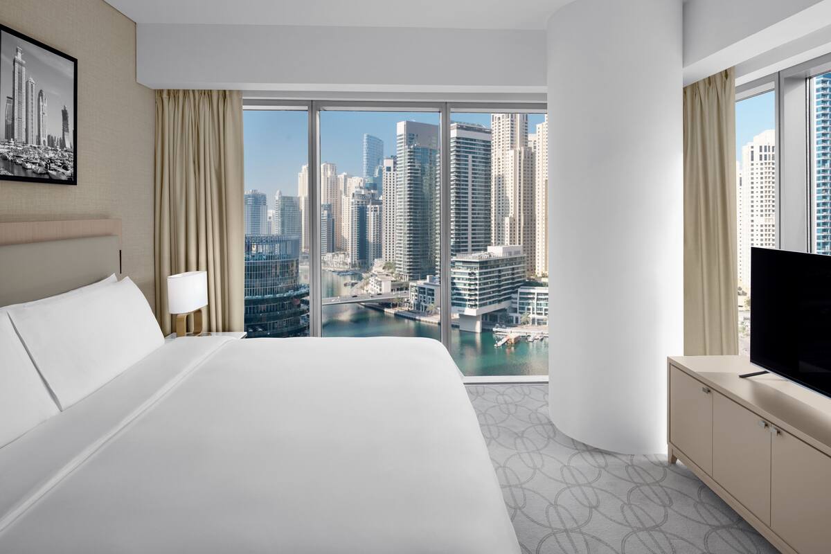 Quarto de hotel com uma cama grande, televisão, escrivaninha e vista para a paisagem urbana através de grandes janelas.