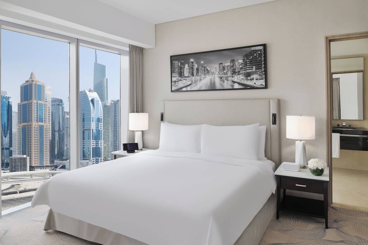 Quarto de hotel com uma cama grande, duas mesas de cabeceira com abajures, vista da cidade através de janelas amplas e uma pintura da cidade emoldurada na parede.