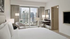 Quarto de hotel com uma cama grande, uma escrivaninha, duas cadeiras, uma TV e vista para edifícios altos.