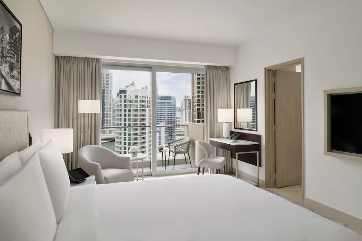 Quarto de hotel com uma cama grande, uma escrivaninha, duas cadeiras, uma TV e vista para edifícios altos.