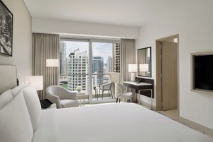 Quarto de hotel com uma cama grande, uma escrivaninha, duas cadeiras, uma TV e vista para edifícios altos.
