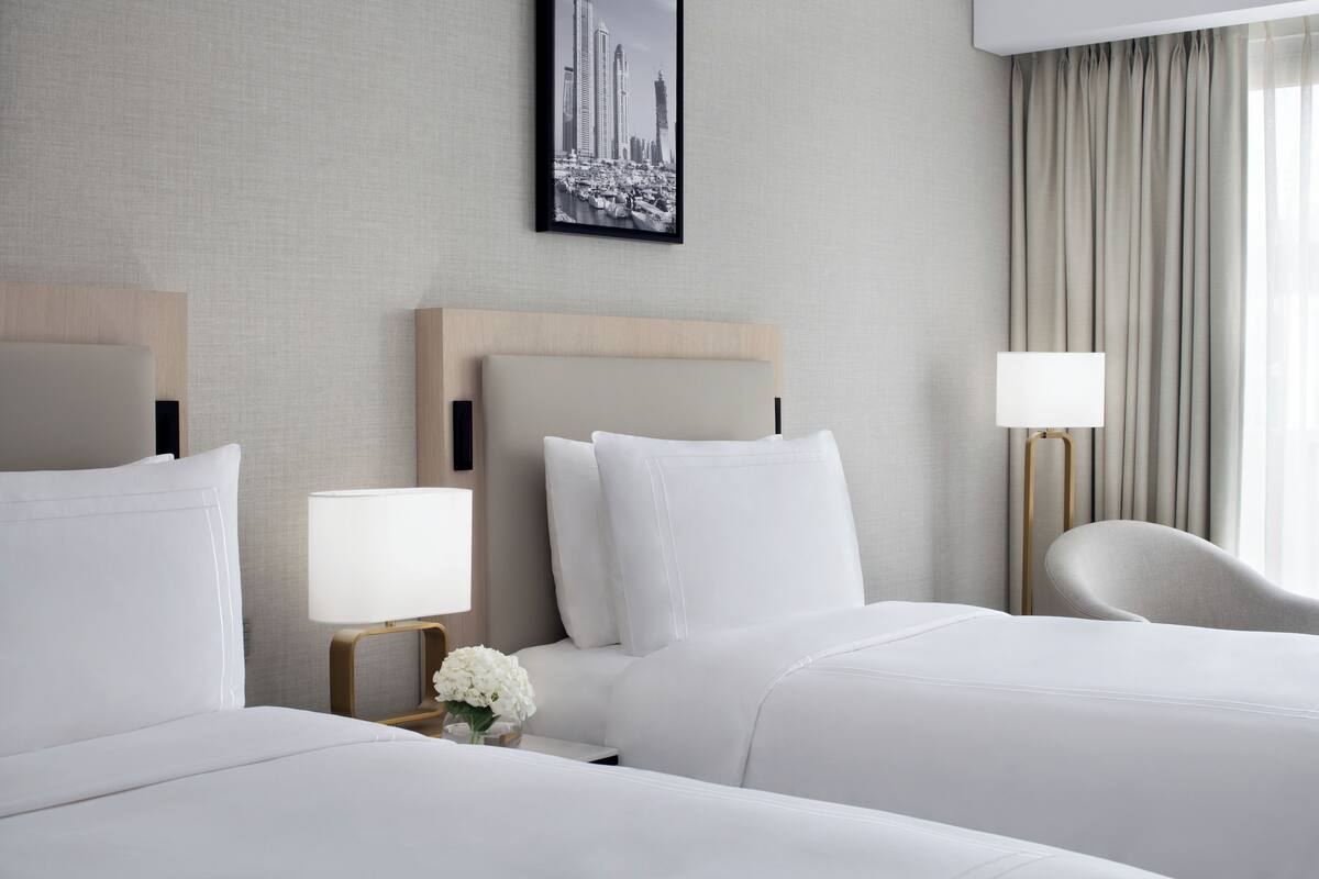 Quarto de hotel com duas camas, uma mesinha de cabeceira com abajur, uma cadeira e uma imagem emoldurada da paisagem urbana na parede.