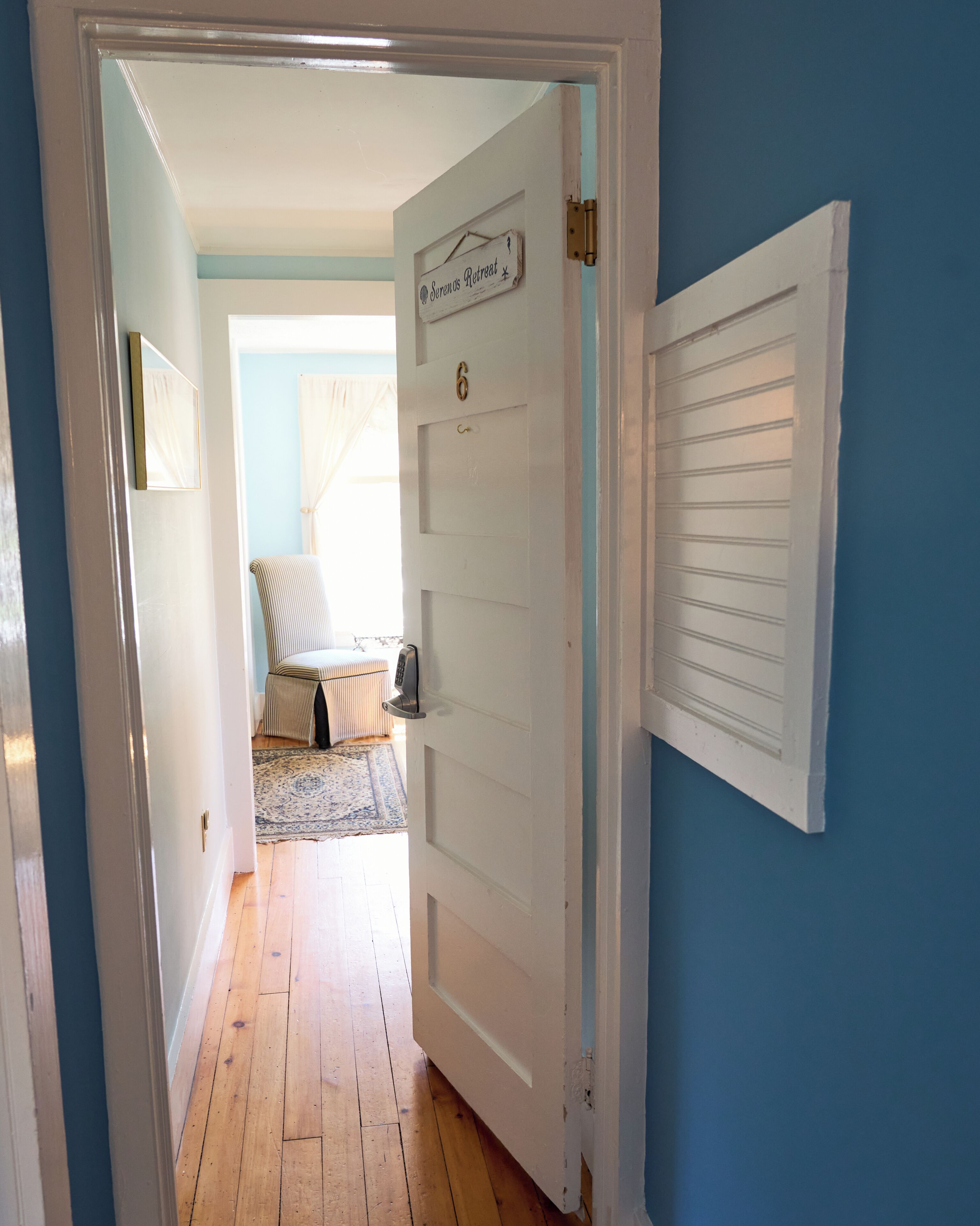 Um corredor com piso de madeira, uma cama com colcha listrada, uma placa com a inscrição 'Serena's Retreat' e uma porta com o número '6'.