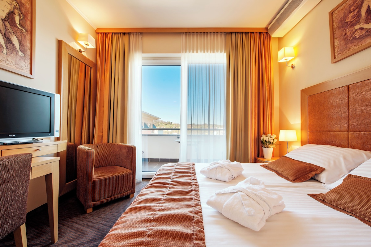 Quarto de hotel com uma cama, uma mesa com televisão, uma cadeira e vista para o exterior através de uma grande janela.