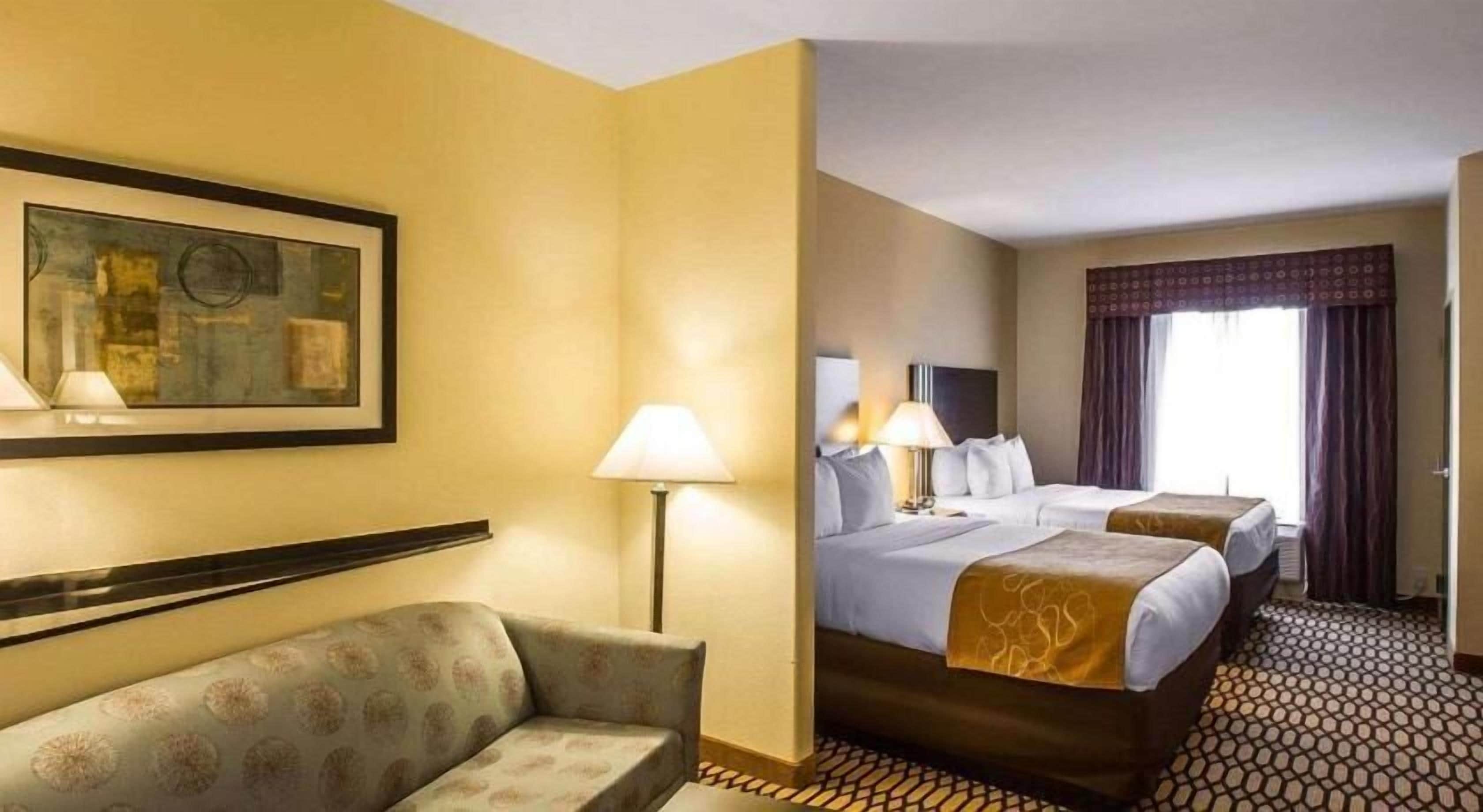 Quarto de hotel com sofá, duas camas, abajur, quadro e piso estampado.