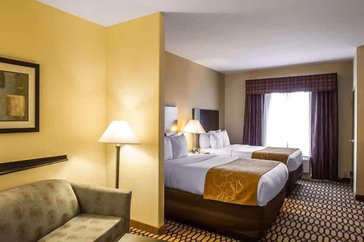 Quarto de hotel com sofá, duas camas, abajur, quadro e piso estampado.