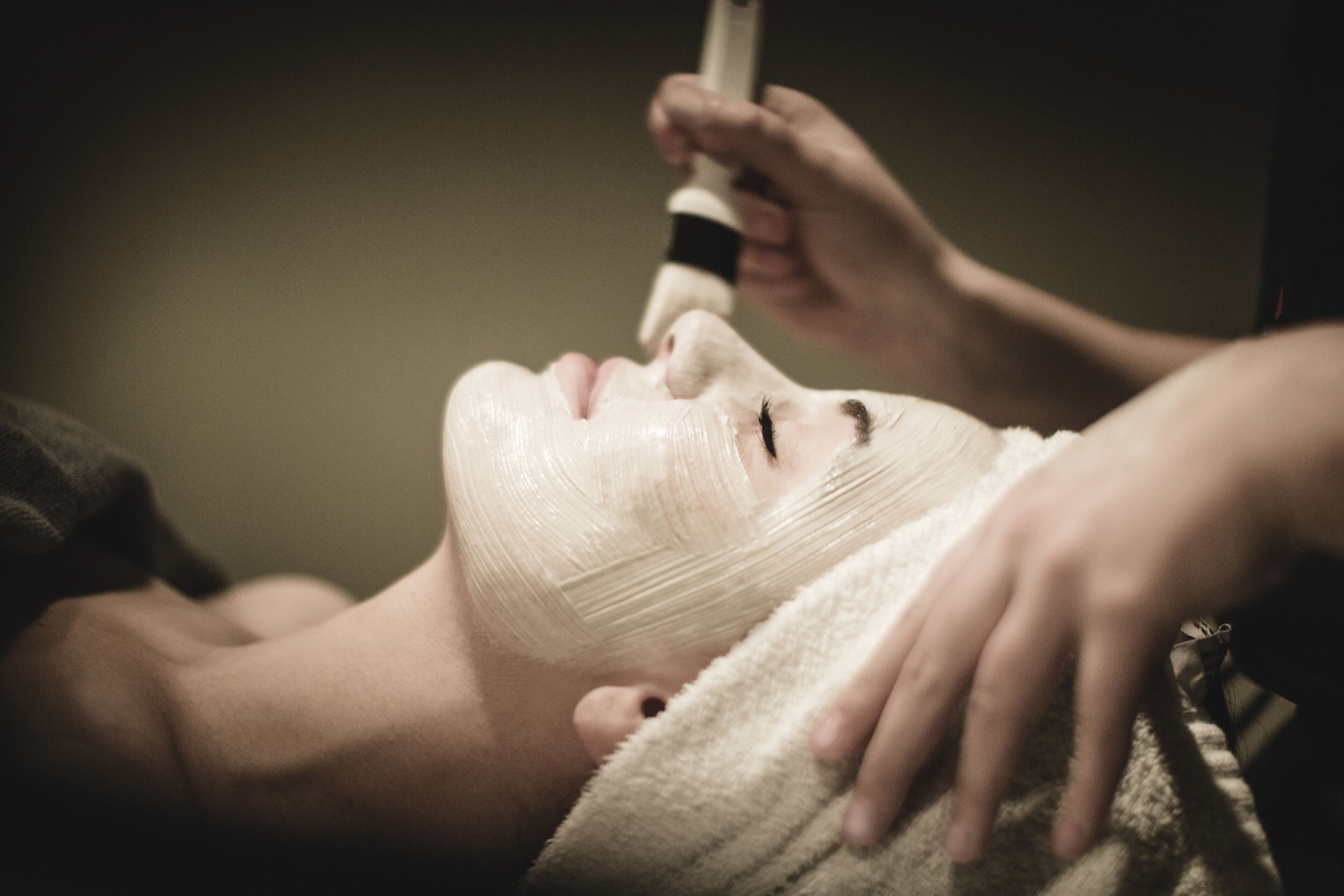 Uma pessoa recebendo tratamento facial com máscara branca, mãos segurando delicadamente a máscara e uma toalha na cabeça.