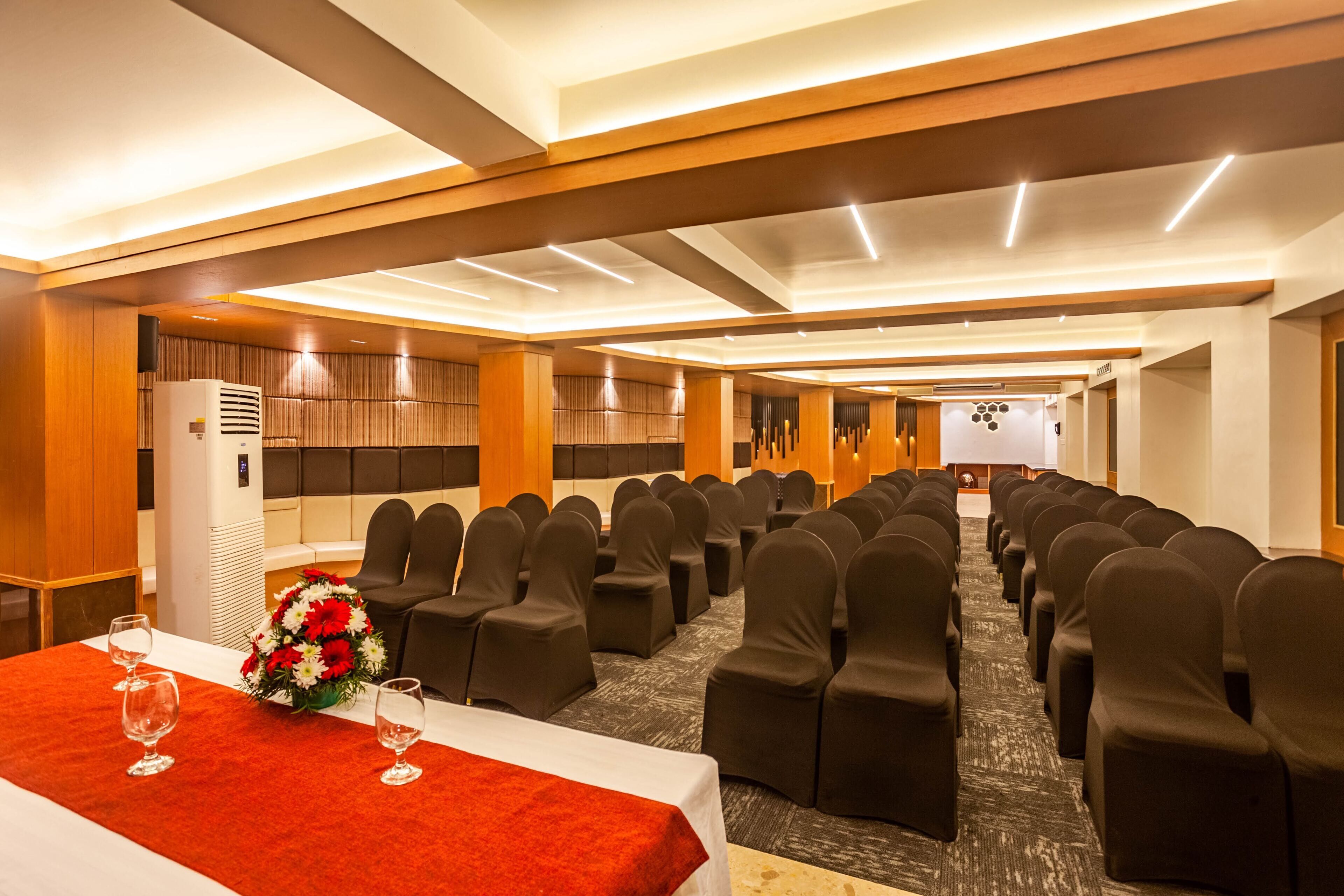 Uma sala de conferências de hotel com fileiras de cadeiras, uma mesa com um jogo americano vermelho e copos, e um arranjo floral.