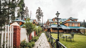 Uma trilha leva a um prédio de madeira com telhado verde, rodeado por árvores e um pequeno gramado com área de estar externa.