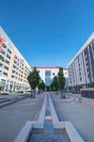 Uma praça urbana moderna com um espelho d'água central, rodeada por edifícios com varandas e um prédio de fachada vermelha e branca em destaque.