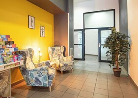 Uma área de recepção com parede amarela, dois poltronas estampadas, uma mesinha com abajur e um vaso de planta.
