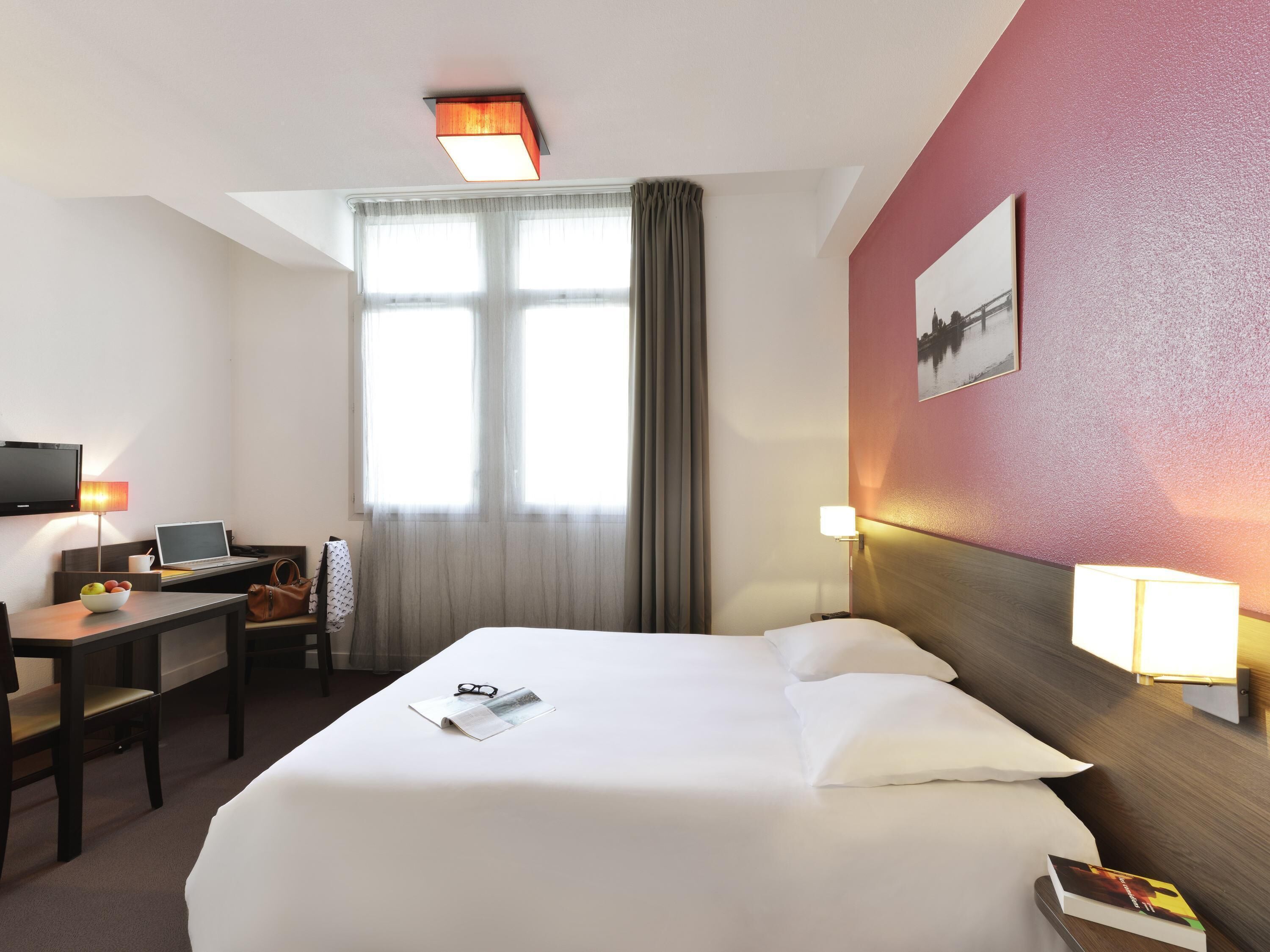 Quarto de hotel com cama, escrivaninha, cadeira, televisão e janela com cortinas.