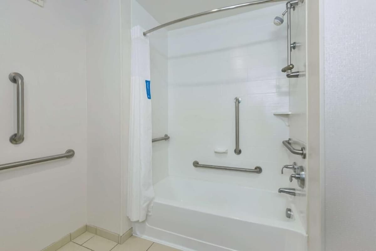 Casa de banho com uma banheira branca, chuveiro com porta de vidro e barras de apoio.