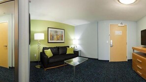 Quarto de hotel com sofá preto, mesa de centro de vidro, luminária, pintura na parede e televisão sobre um suporte de madeira.