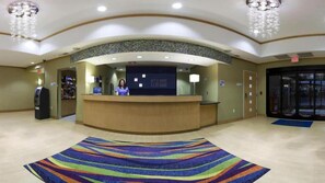 Uma área de recepção de hotel com um balcão em arco, um recepcionista, um quiosque de autoatendimento e um tapete listrado e colorido.