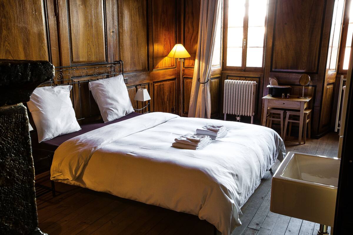 Quarto de hotel com uma cama grande, uma mesa de cabeceira com luminária, um radiador e uma pequena escrivaninha com espelho.