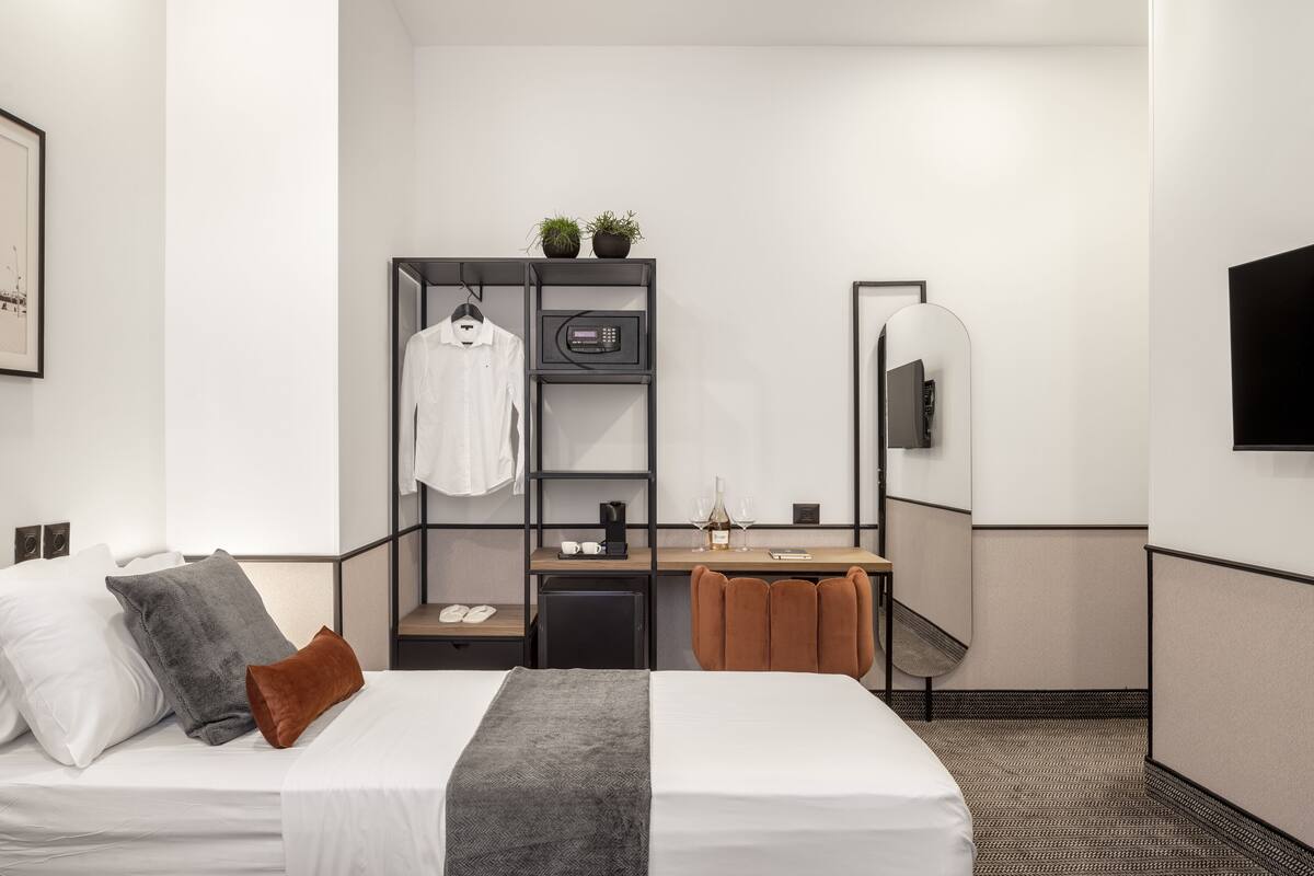 Quarto de hotel moderno com uma cama bem arrumada, uma escrivaninha com computador, uma pequena área de estar e uma TV fixada na parede.