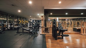 Academia com diversos equipamentos de exercício, piso de madeira e uma área com estantes de livros.