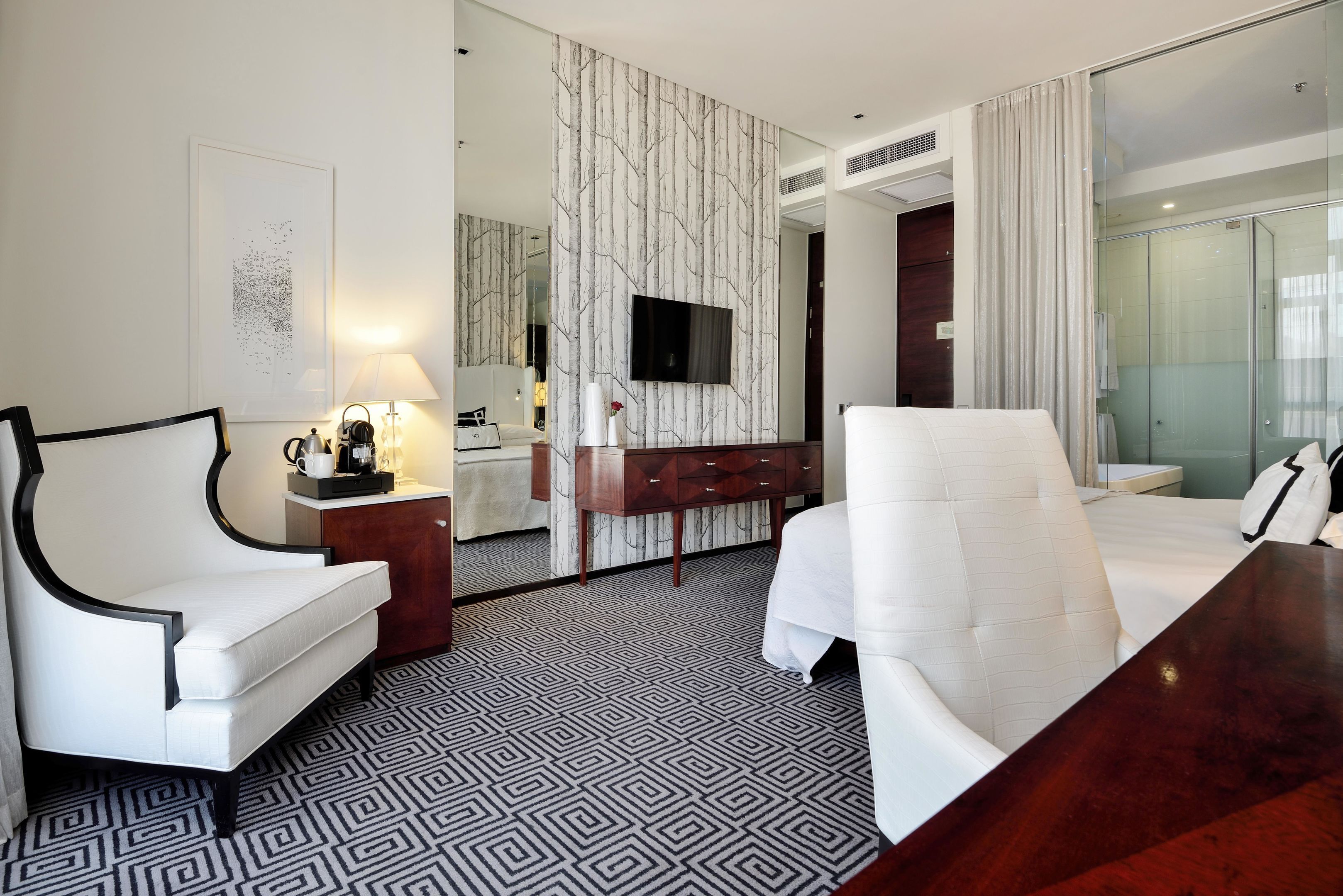 Quarto de hotel moderno com uma cama grande, duas poltronas brancas, uma escrivaninha de madeira, uma TV fixada na parede e um chão estampado.