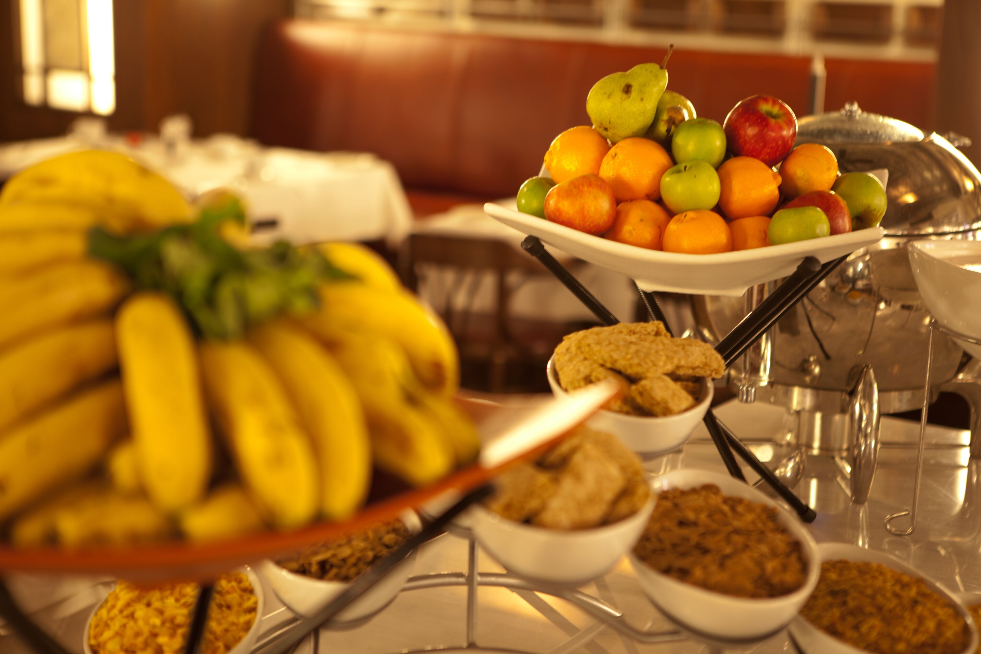 Uma mesa de buffet com diversos alimentos, incluindo bananas, laranjas, maçãs e cereais.