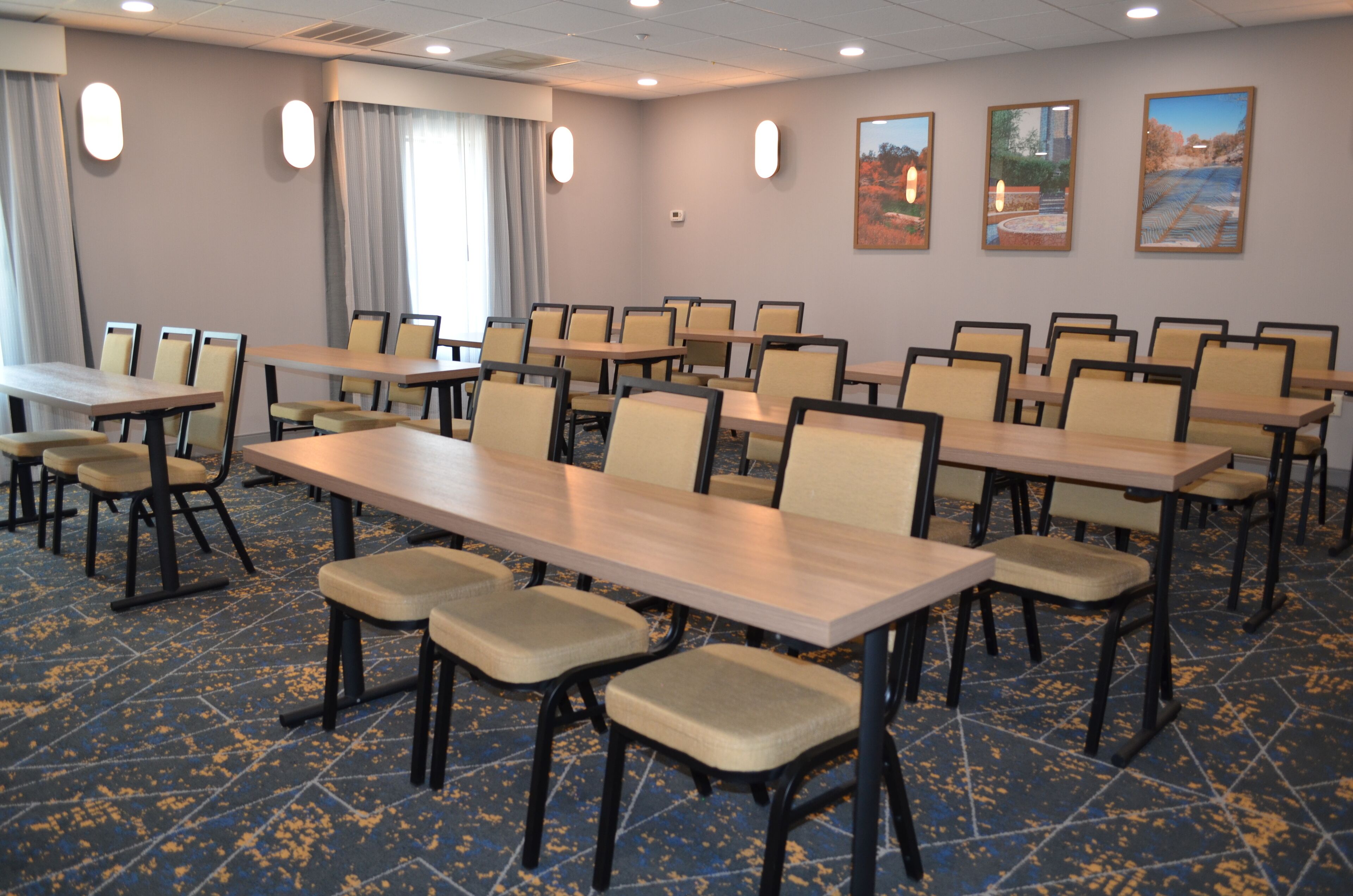 Uma sala de conferências com várias mesas e cadeiras, janelas amplas com cortinas e quadros decorativos nas paredes.