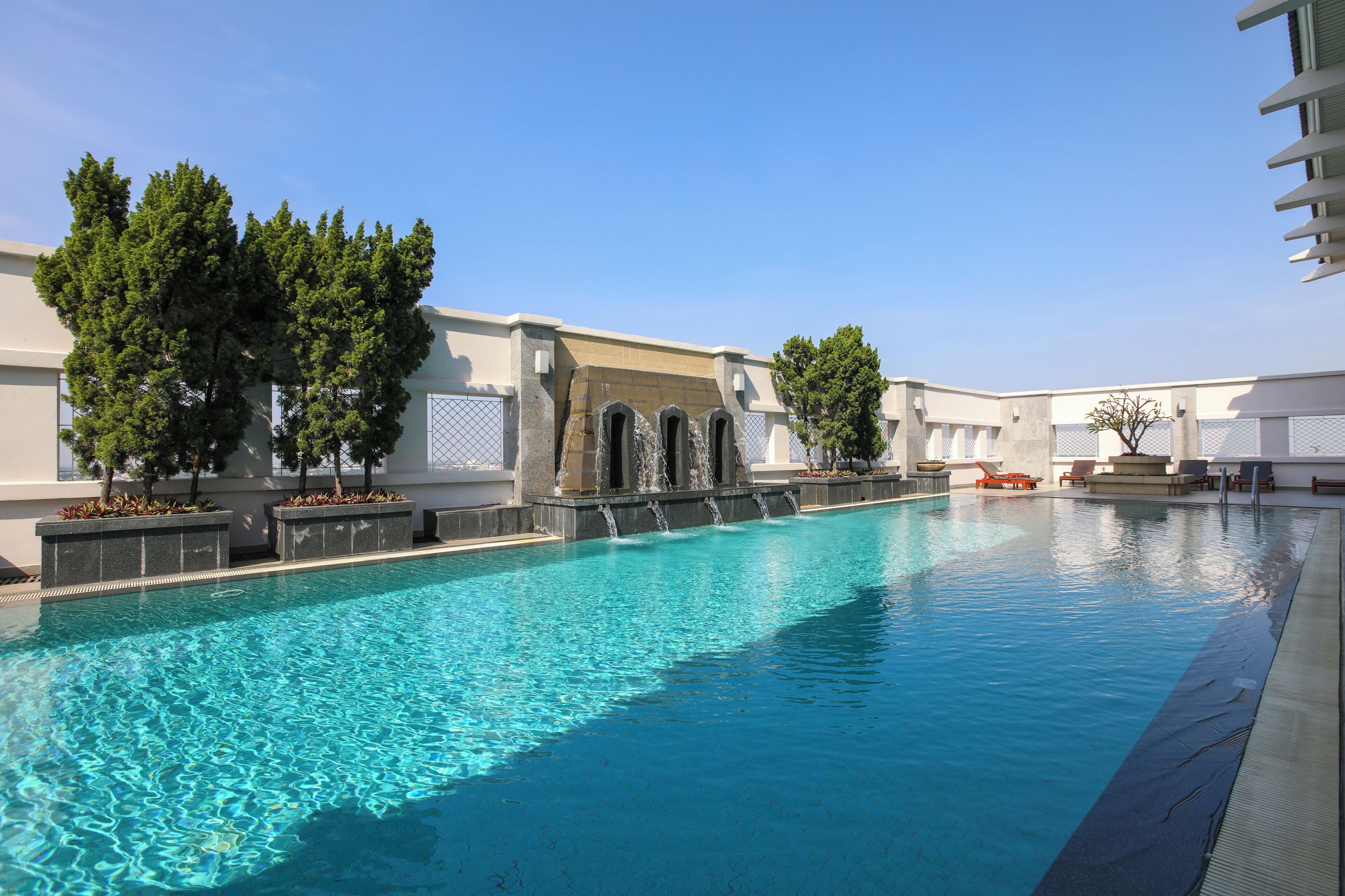 Piscina com água azul cristalina, cercada por um edifício moderno com fachada em azulejos e algumas árvores.