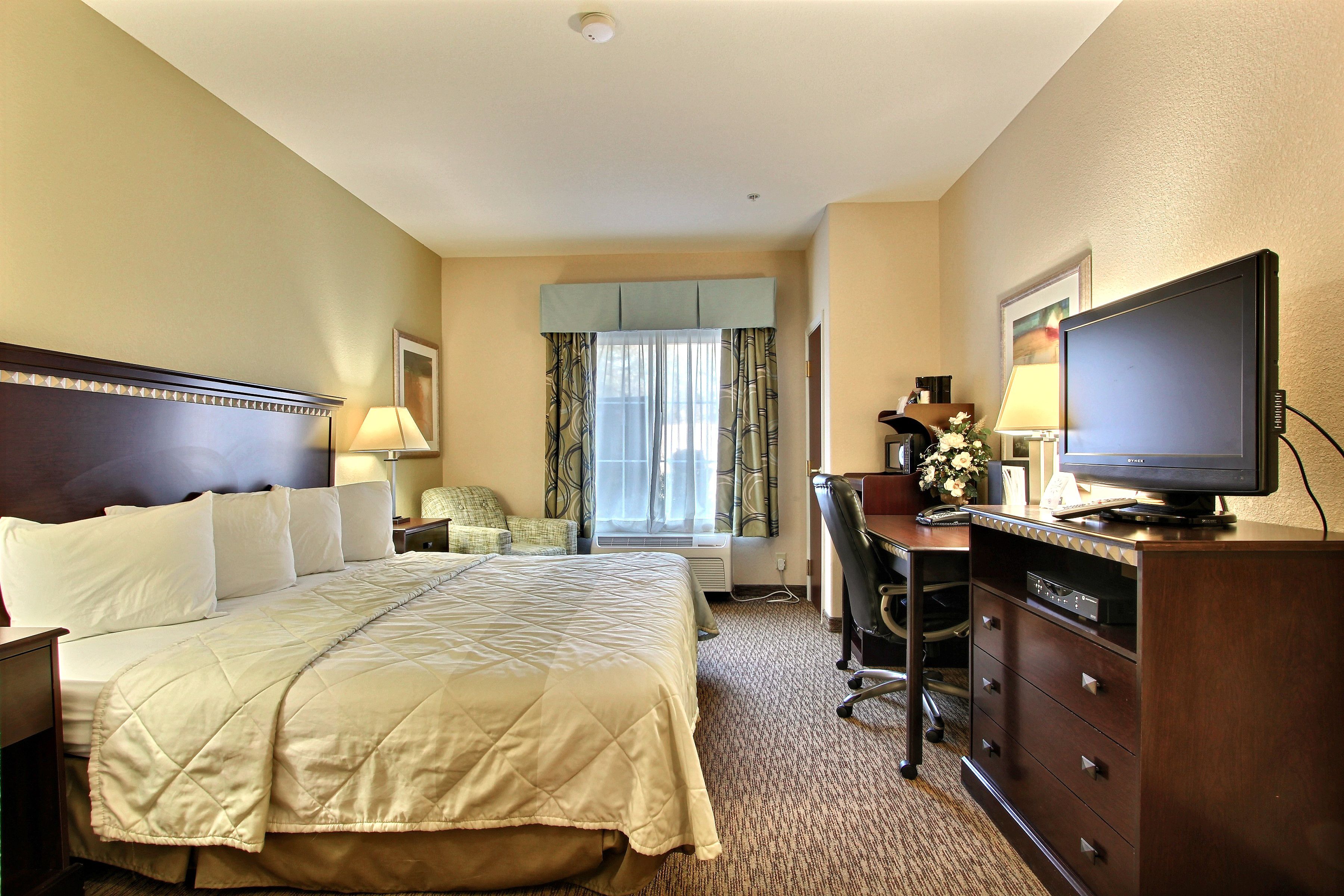 Quarto de hotel com uma cama grande, uma escrivaninha com televisão, uma cadeira e um abajur.