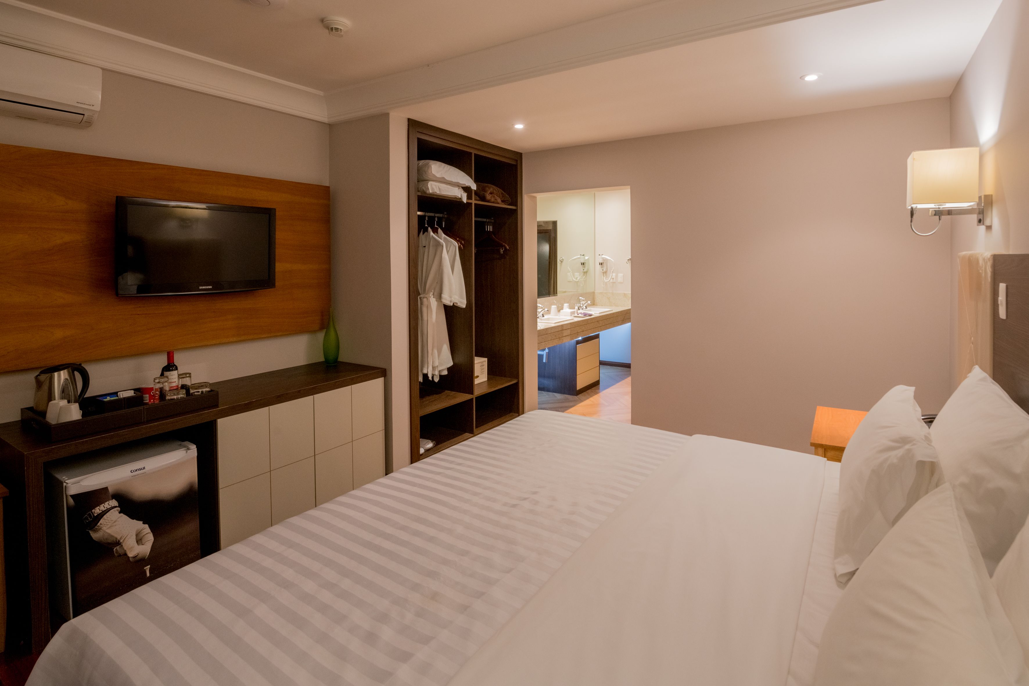 Quarto de hotel com cama, televisão na parede, guarda-roupa, secretária com cafeteira e casa de banho visível por uma porta aberta.