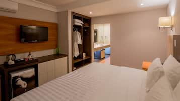Quarto de hotel com cama, televisão na parede, guarda-roupa, secretária com cafeteira e casa de banho visível por uma porta aberta.