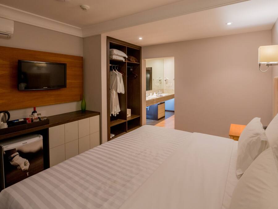 Quarto de hotel com cama, televisão na parede, guarda-roupa, secretária com cafeteira e casa de banho visível por uma porta aberta.